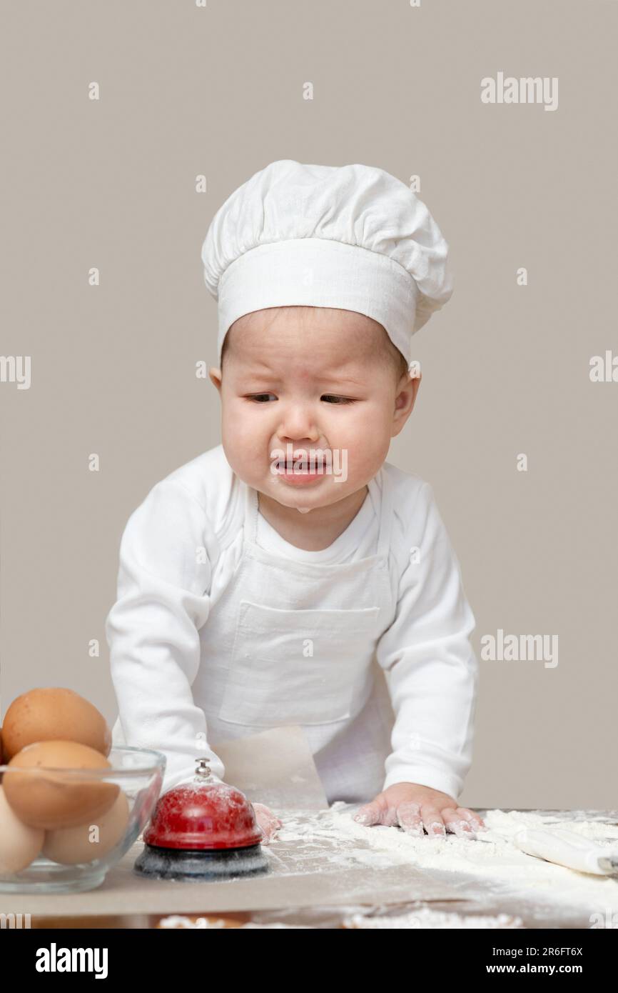 An Asian baby in a chef's cap and apron, smeared in flour, prepares ...