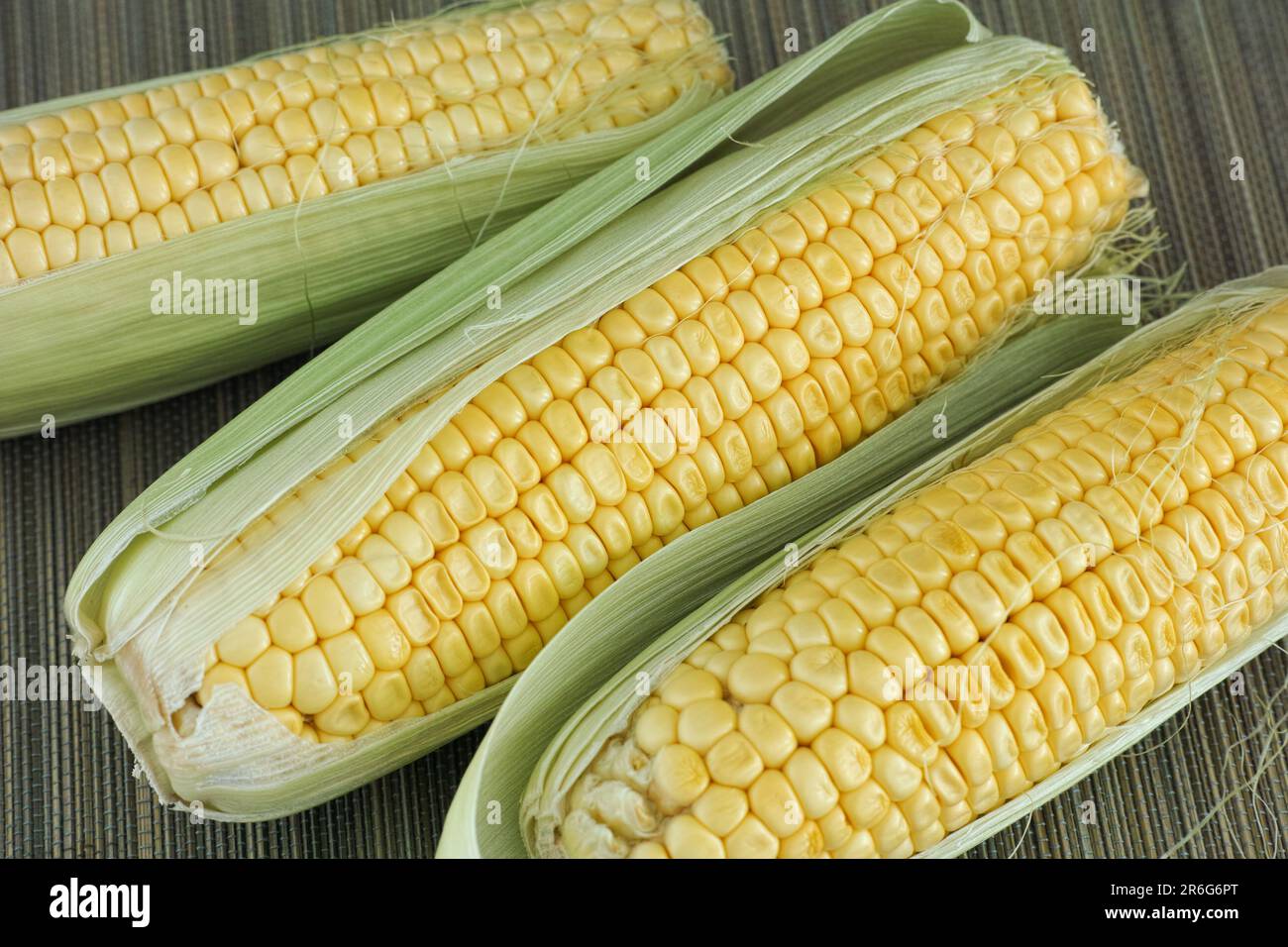 Three corn cobs with the husk lying on a mat. Close up Stock Photo - Alamy