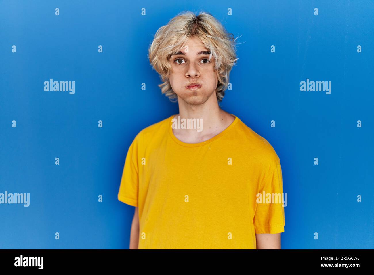 Young modern man standing over blue background puffing cheeks with ...