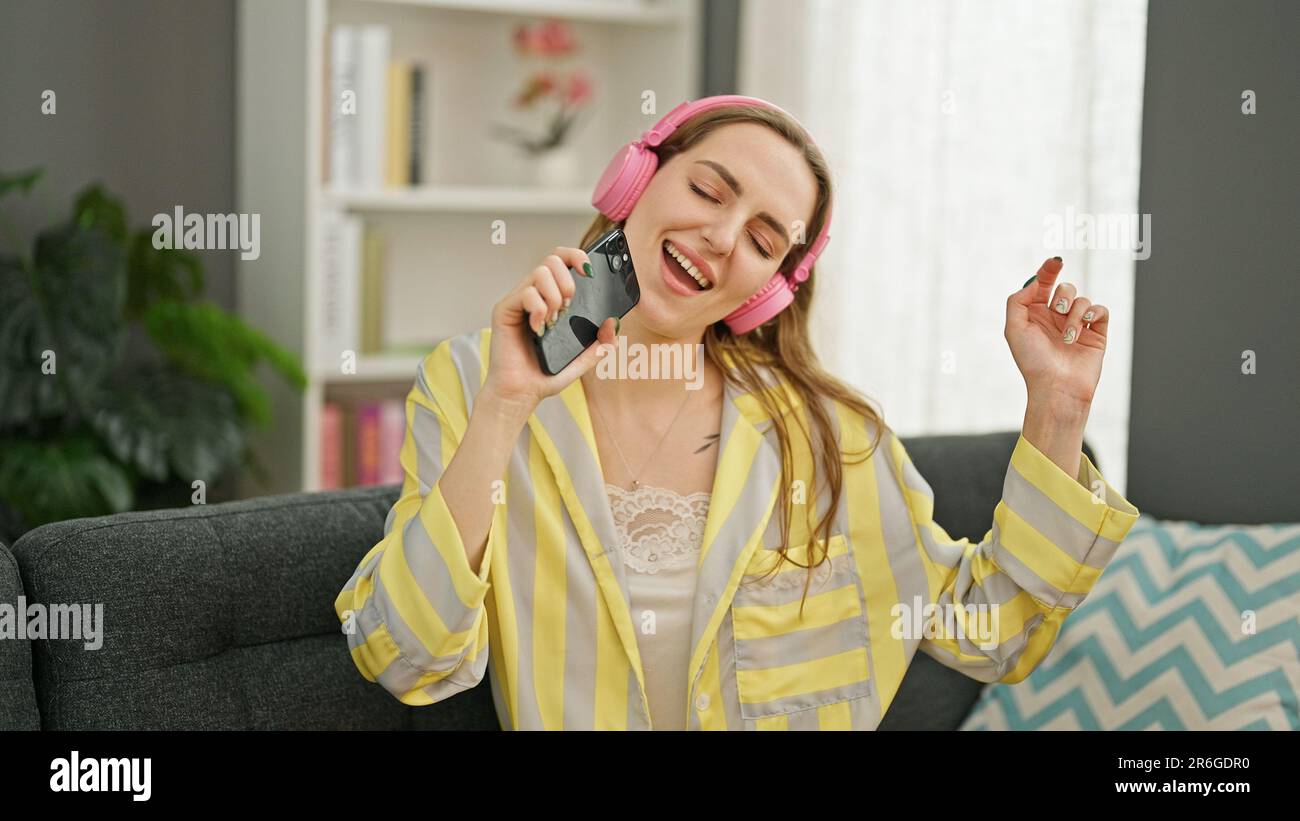 Young blonde woman listening to music sitting on the sofa singing song ...