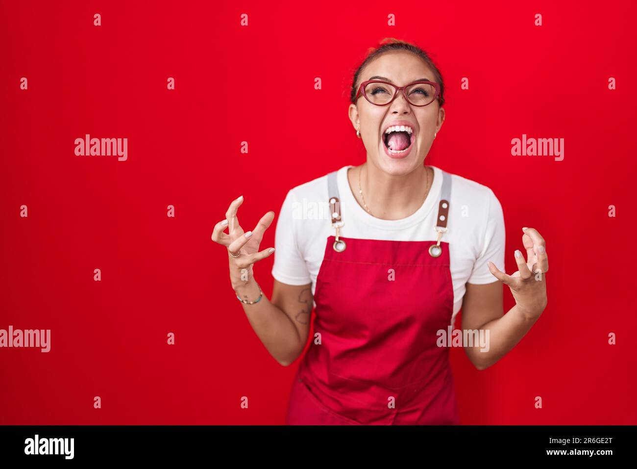 Young hispanic woman wearing waitress apron over red background crazy ...