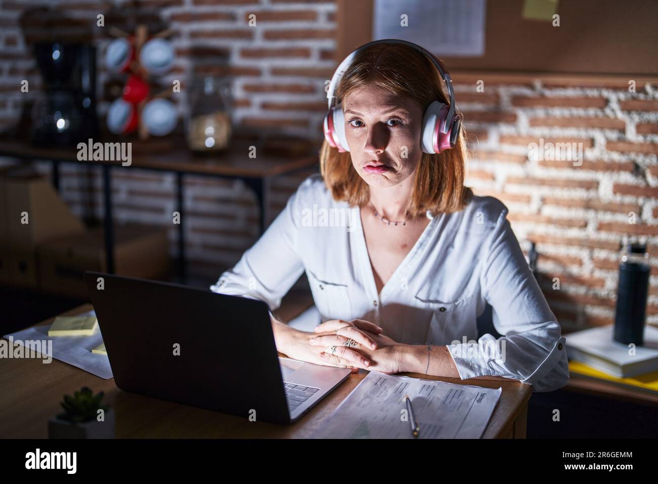 Young redhead woman working at the office at night wearing headphones ...
