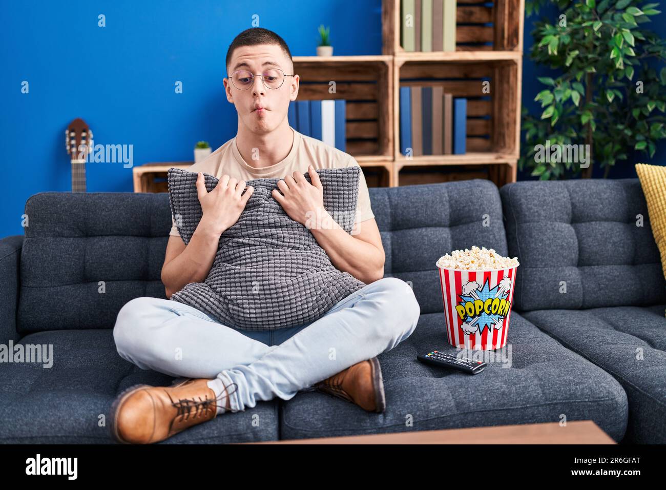 Young man eating popcorn making fish face with mouth and squinting eyes ...