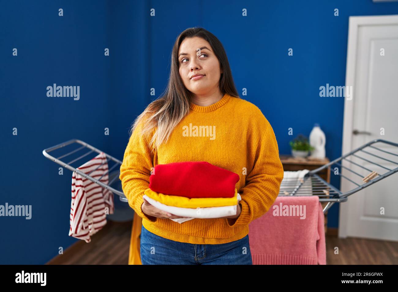 Young hispanic woman holding clean and folded laundry smiling looking to the side and staring ...