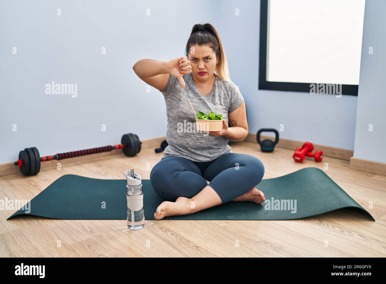 Young hispanic woman sitting on yoga mat eating healthy salad with ...