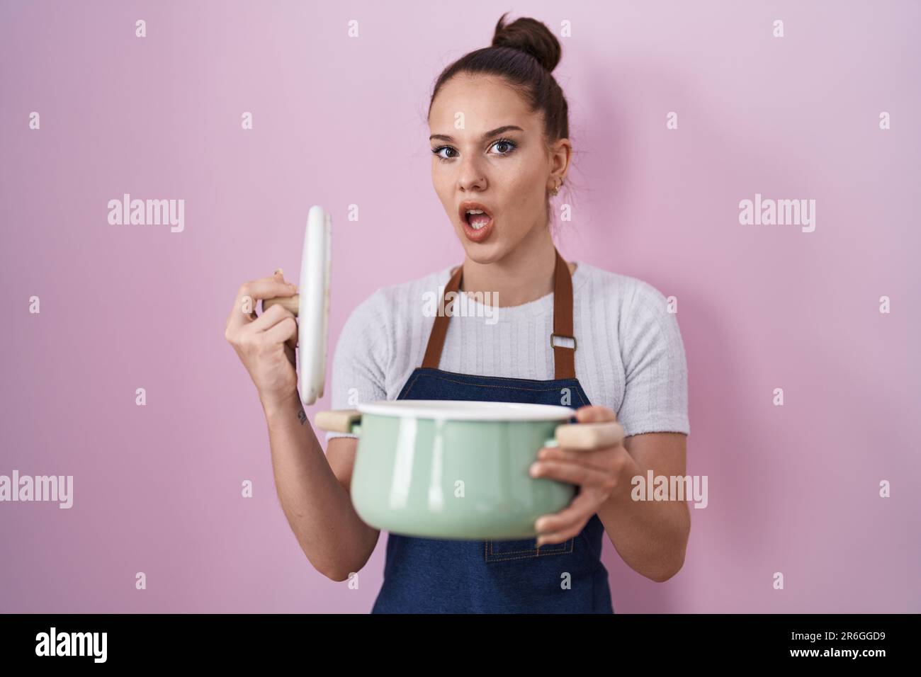 Young hispanic girl wearing apron holding cooking pot in shock face ...