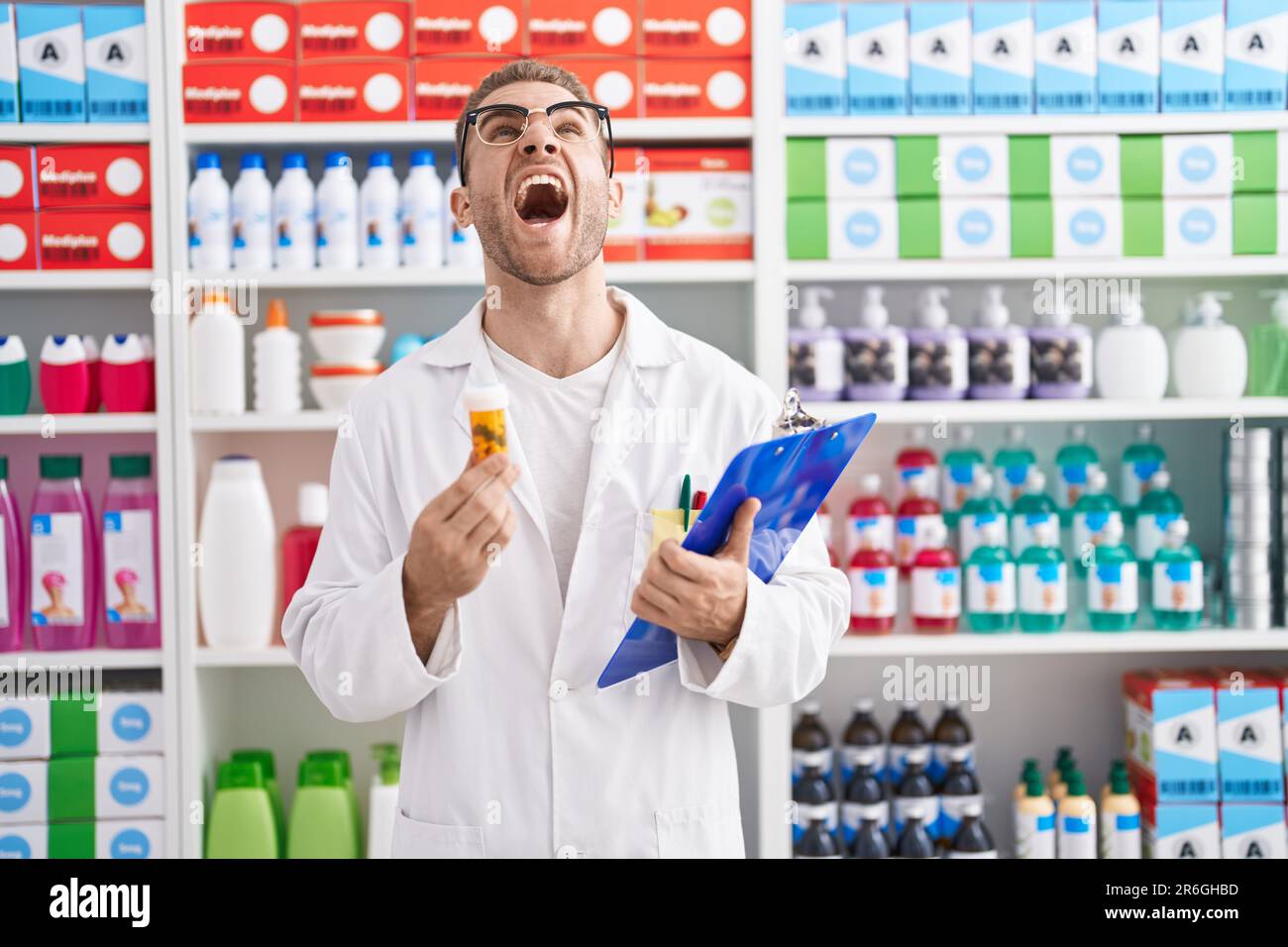 Young caucasian man working at pharmacy drugstore holding pills angry ...
