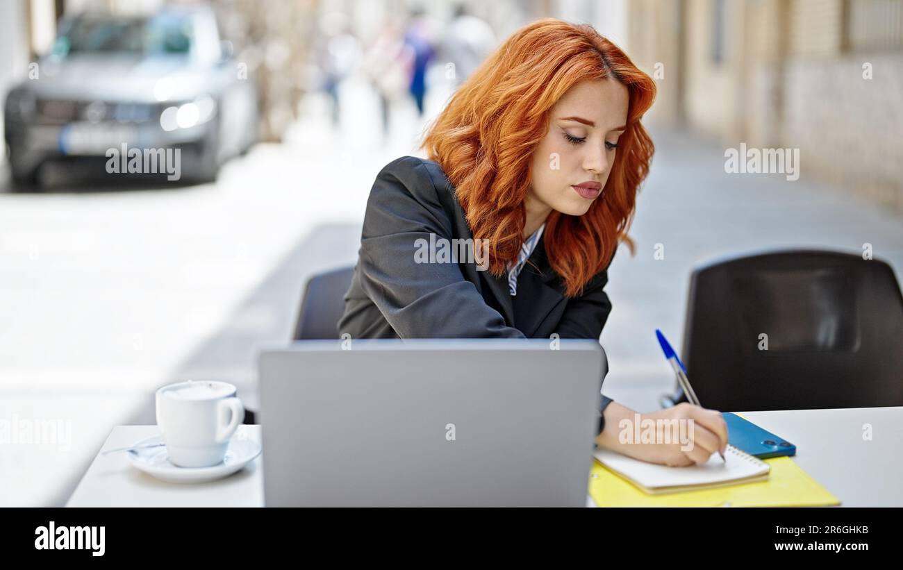 Young redhead woman business worker using laptop taking notes at coffee ...