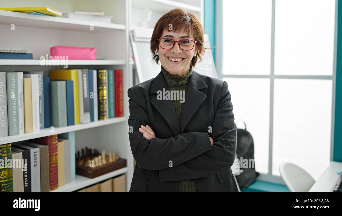 Mature hispanic woman teacher standing by white board with crossed arms at library university ...