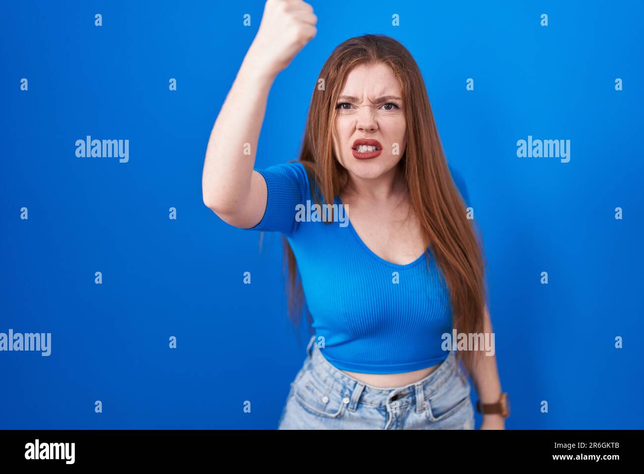 Redhead woman standing over blue background angry and mad raising fist ...