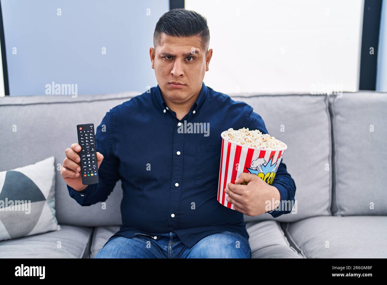 Hispanic young man eating popcorn using tv control skeptic and nervous ...