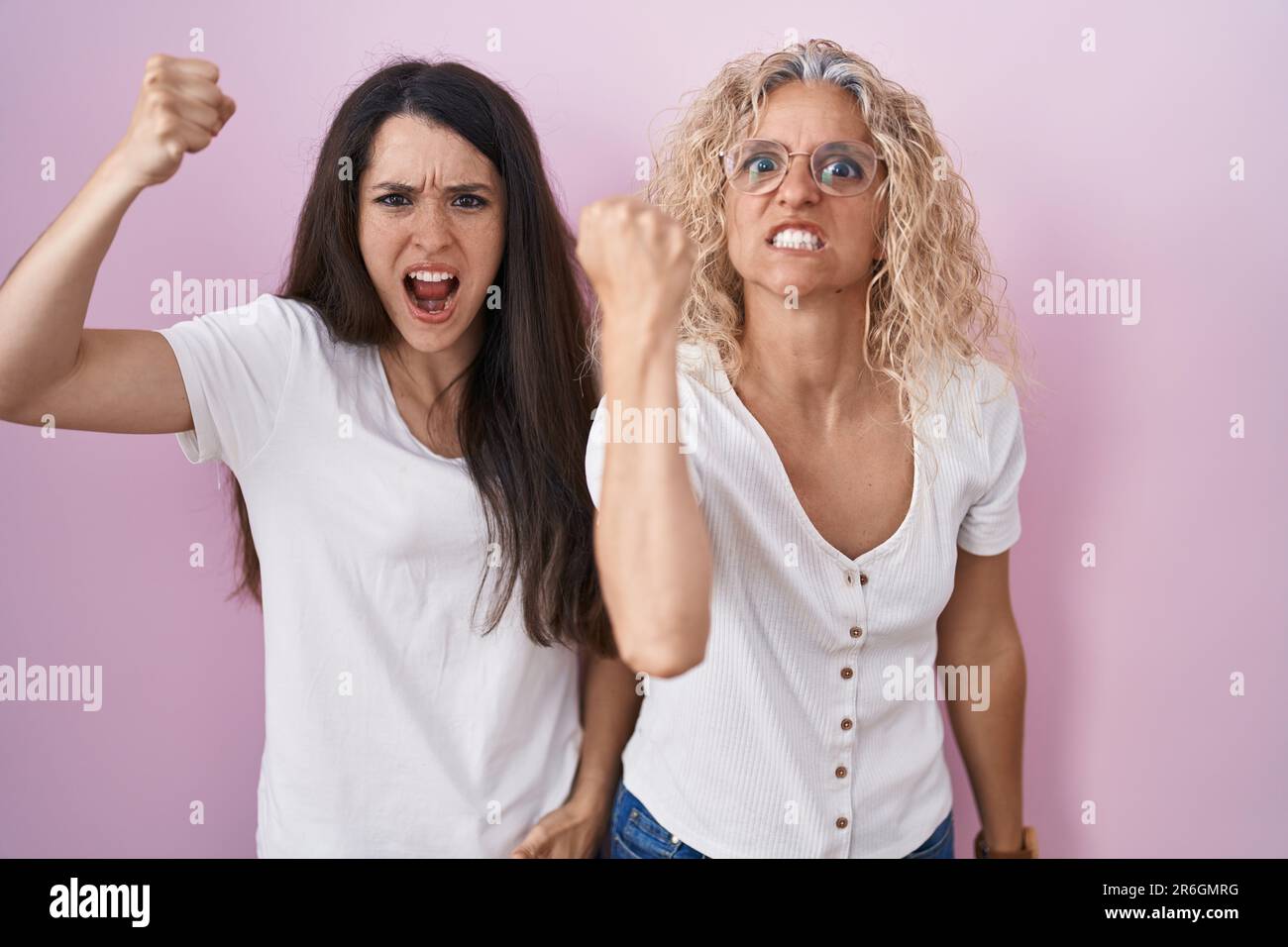 Mother and daughter standing together over pink background angry and ...