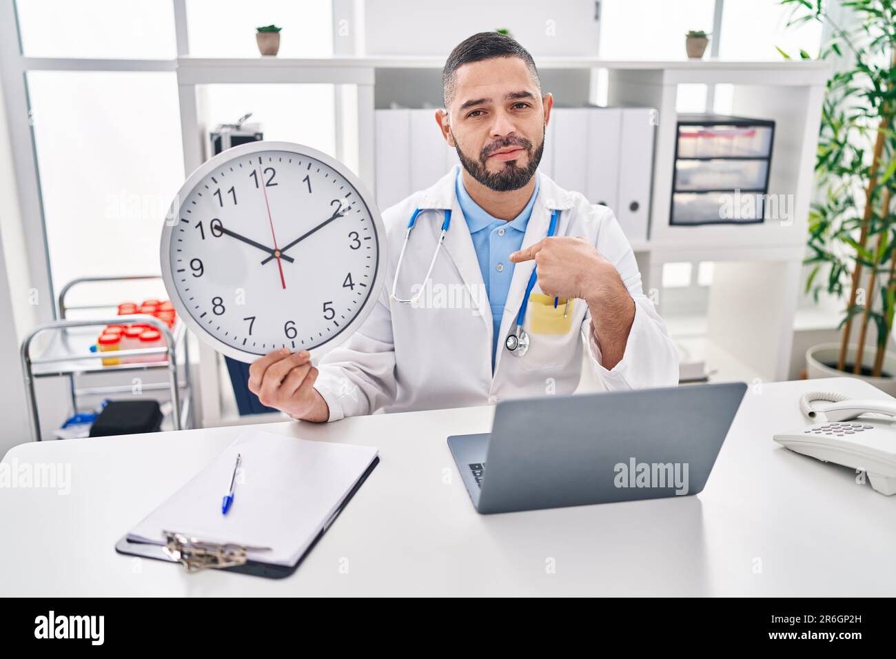 Hispanic doctor man holding clock at the clinic pointing finger to one ...