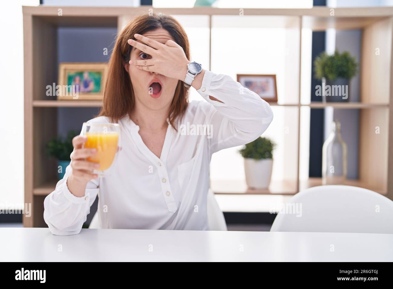 Brunette woman drinking glass of orange juice peeking in shock covering ...
