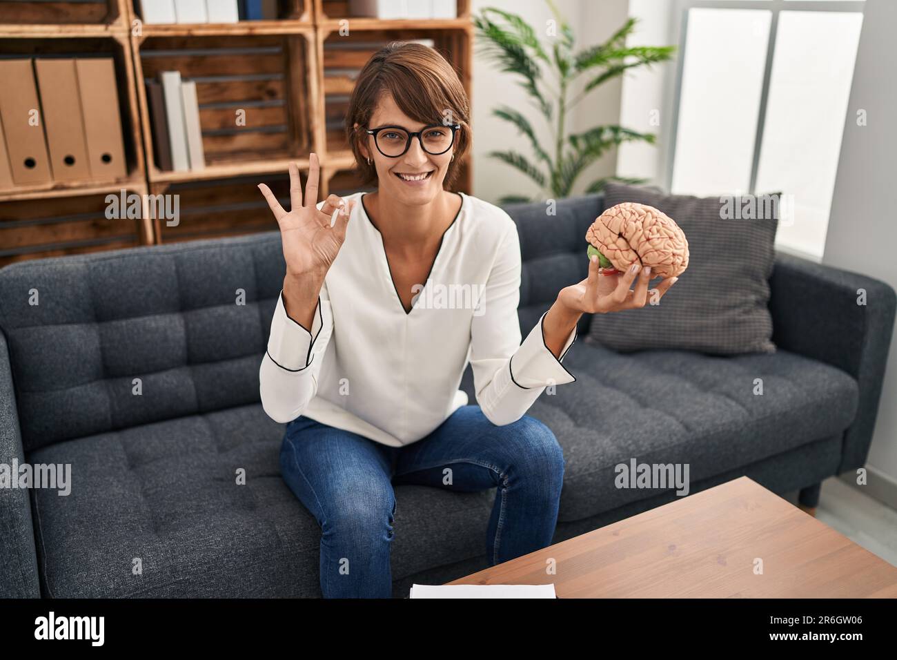 Brunette woman working at therapy office holding brain doing ok sign ...
