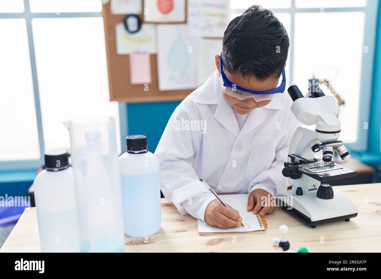 Adorable hispanic boy student using microscope writing notes at ...