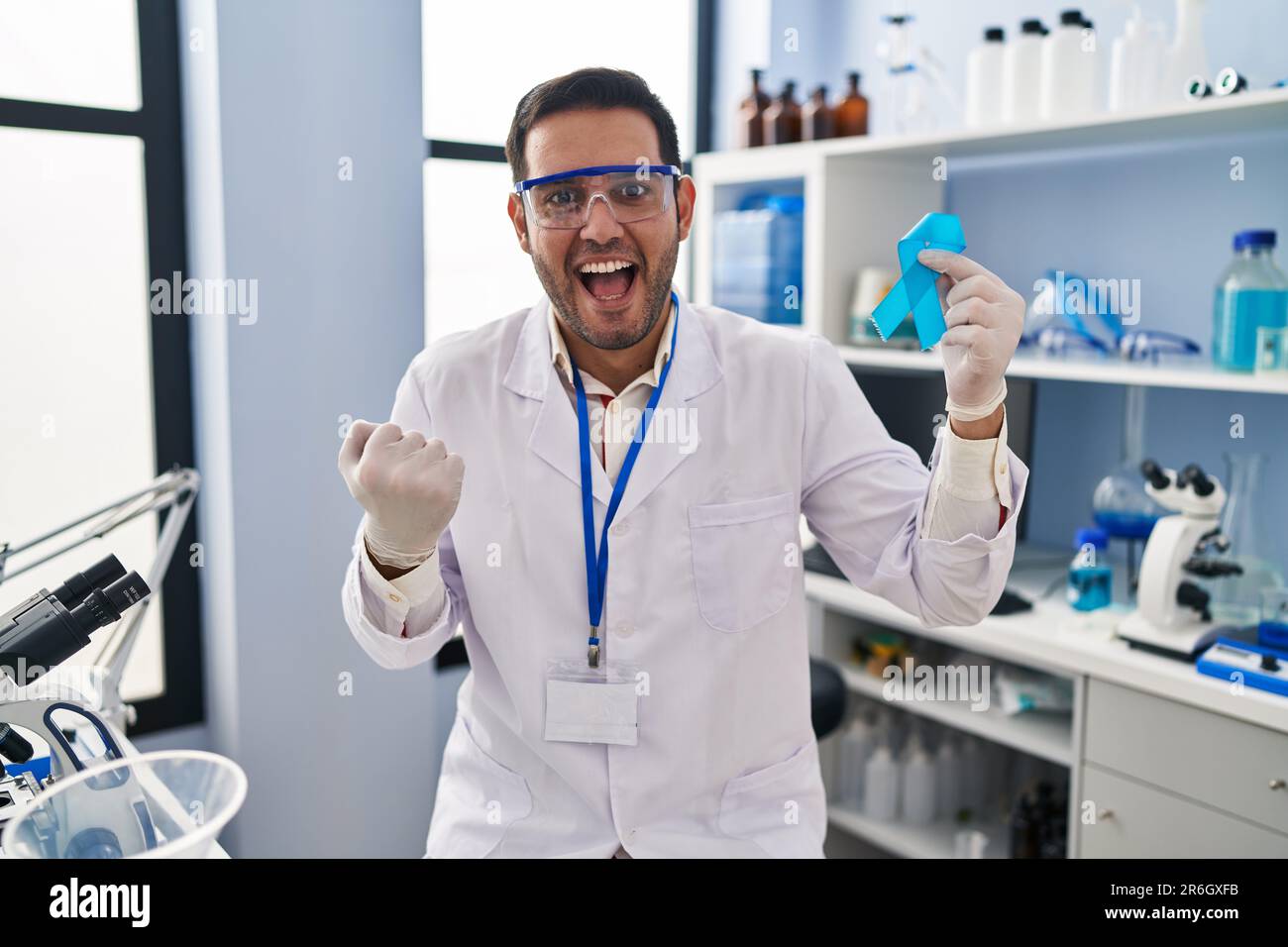 Young hispanic man with beard working at scientist laboratory holding blue ribbon screaming ...