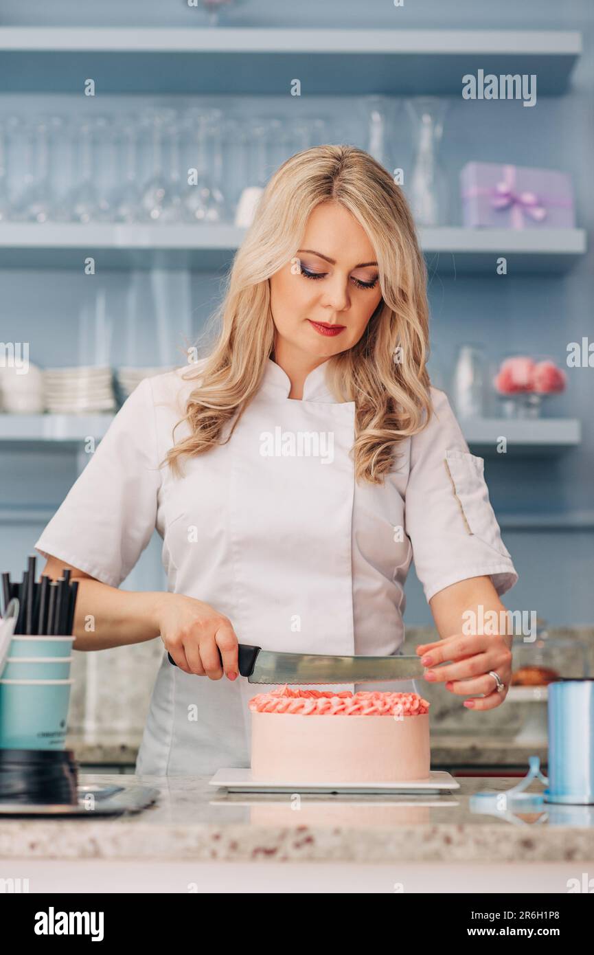 Portrait of cheerful baker woman cut pink cake, standing behind the ...