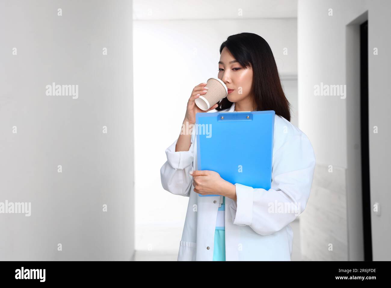 Female Asian medical intern with clipboard drinking coffee in clinic ...