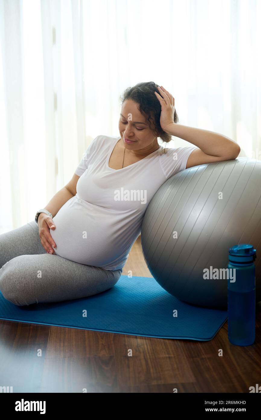 Happy pregnant woman gently stroking belly, relaxing on a yoga mat ...