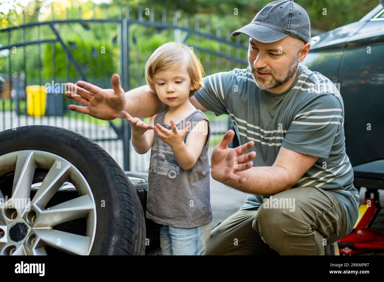 Cute toddler boy helping his father to change car wheels at their backyard. Father teaching his ...