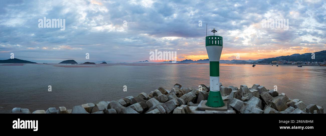 A lighthouse on the stony long pier near the coastal town at sunset ...