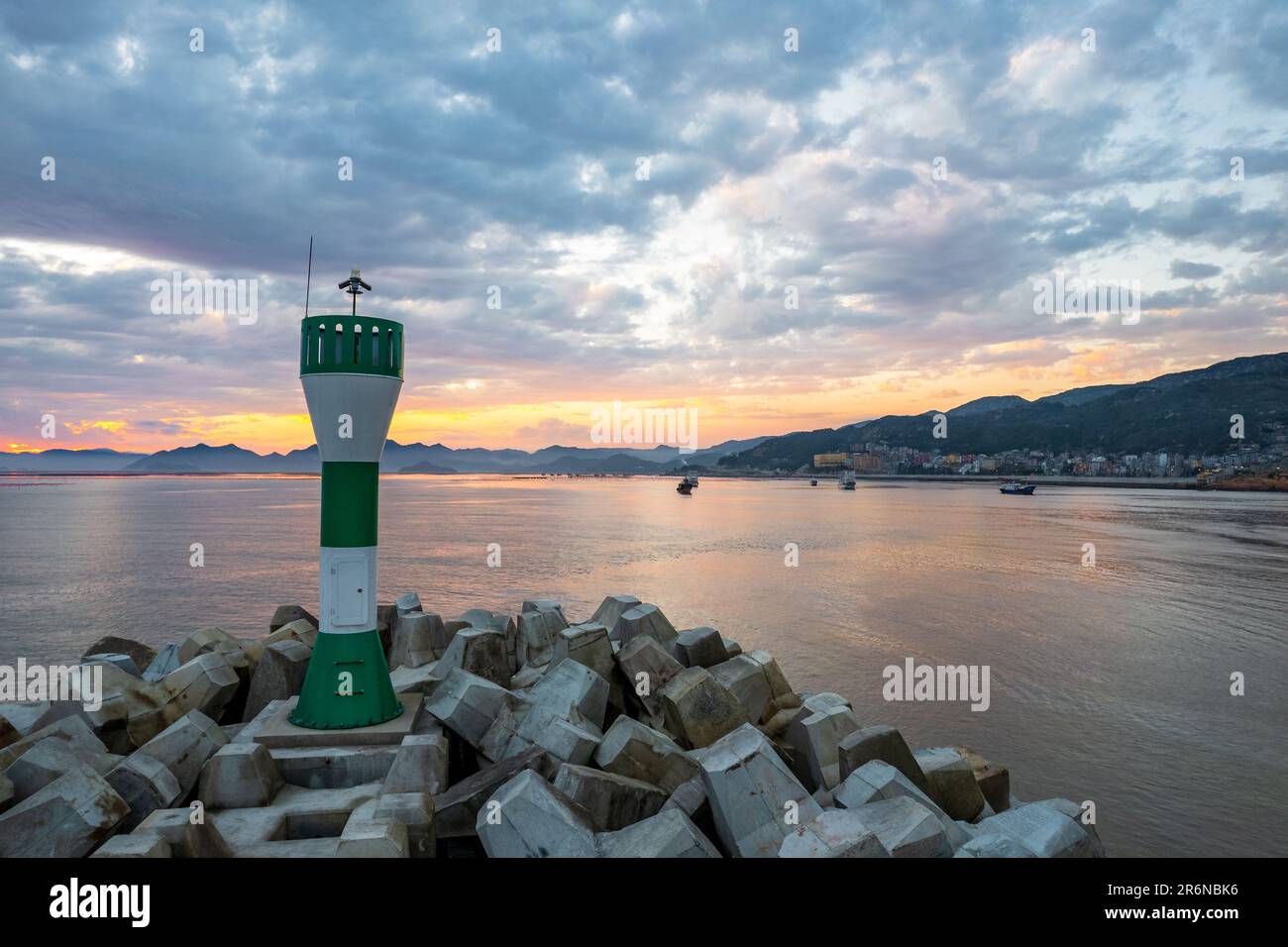 A lighthouse on the stony long pier near the coastal town at sunset ...