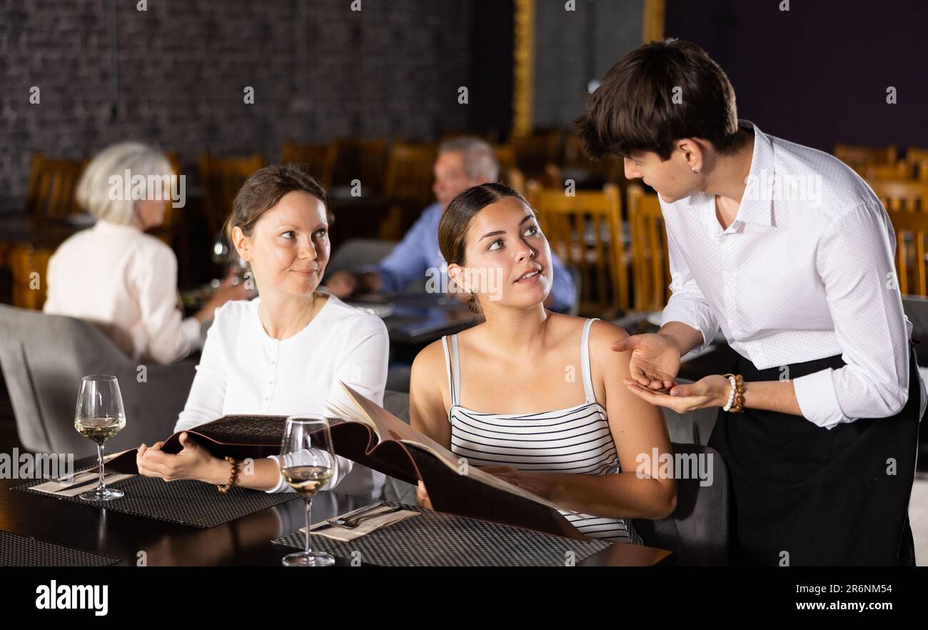 Young waiter recommending dishes from menu to female guests in ...