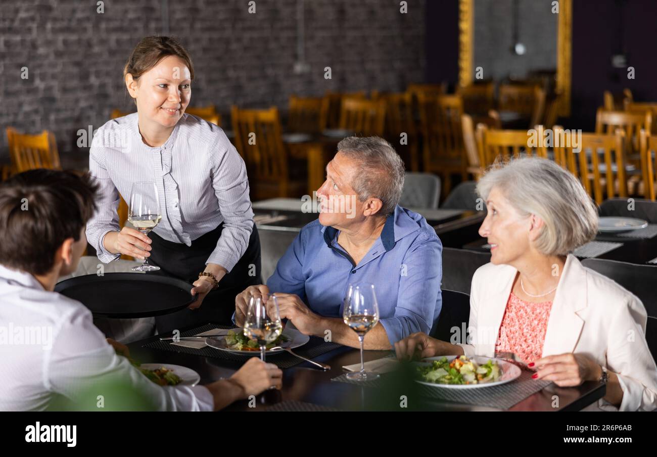 Waitress girl serves glass of wine to elderly couple and adult son ...
