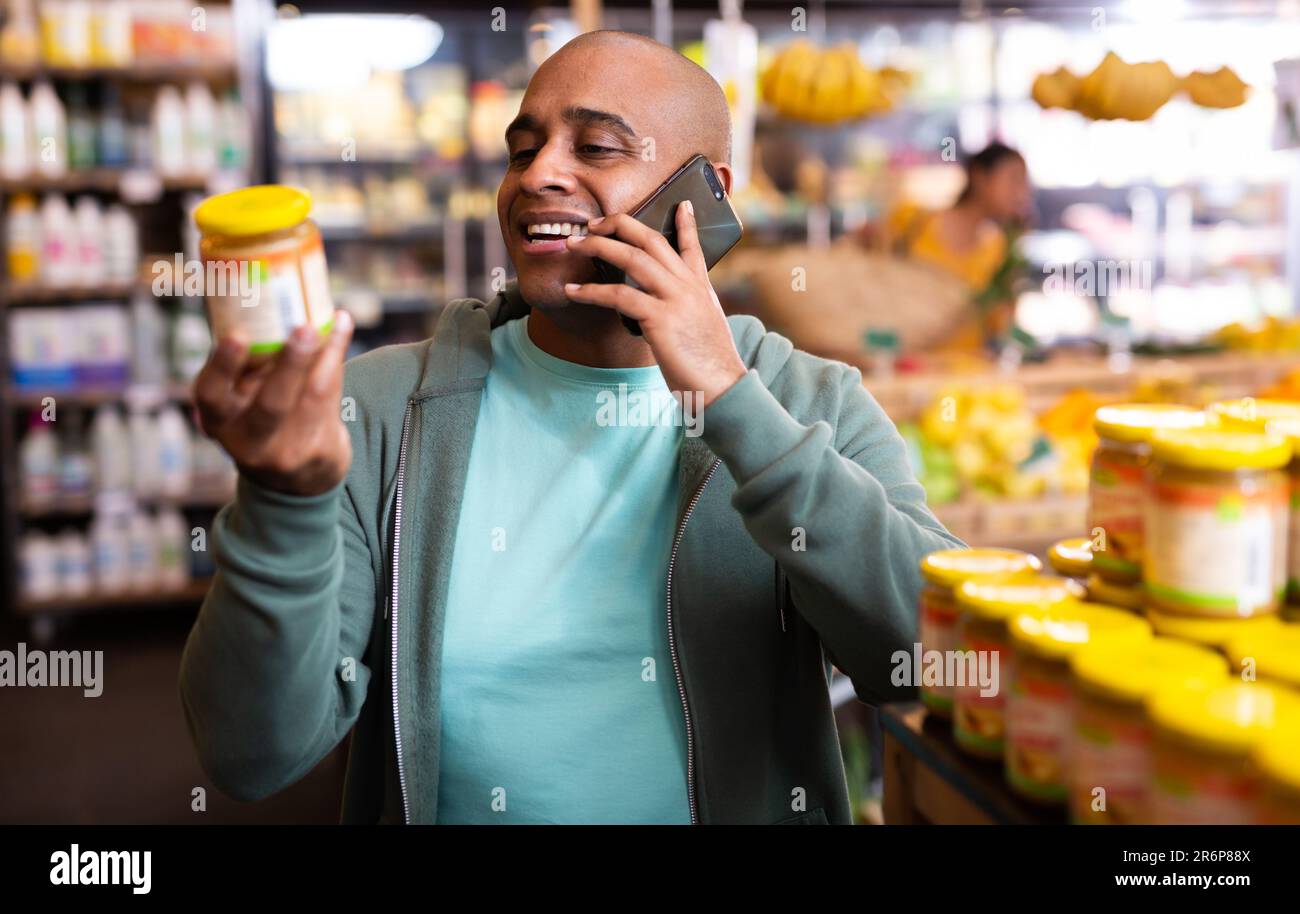 Positive man choosing canned food and talking on mobile phone in ...