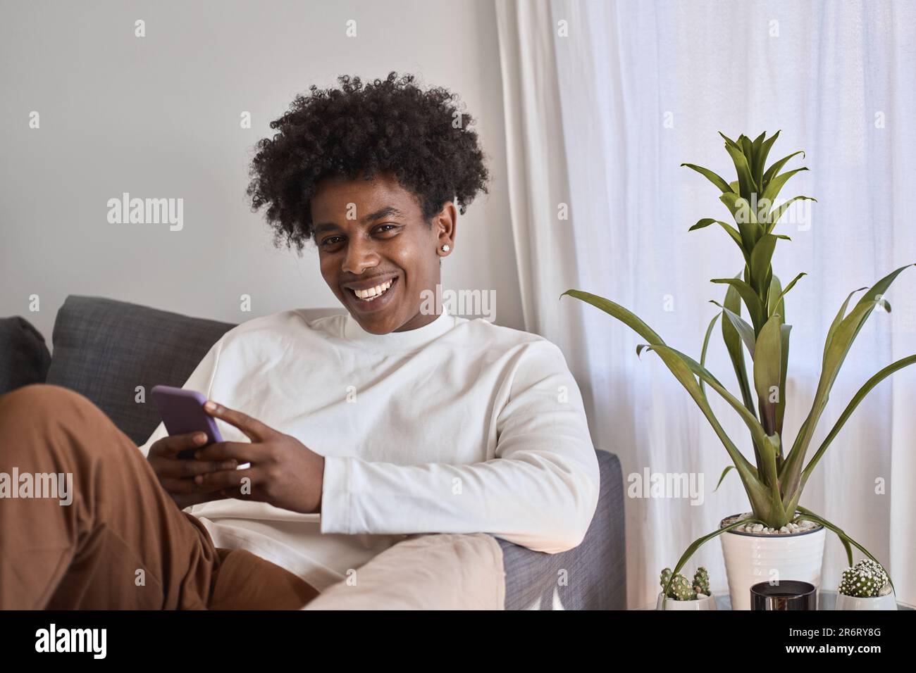 Smiling gen z African American teen sitting on couch using smartphone ...