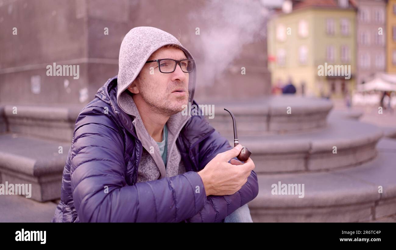 Adult man with glasses sitting on square and smoking a tobacco pipe ...