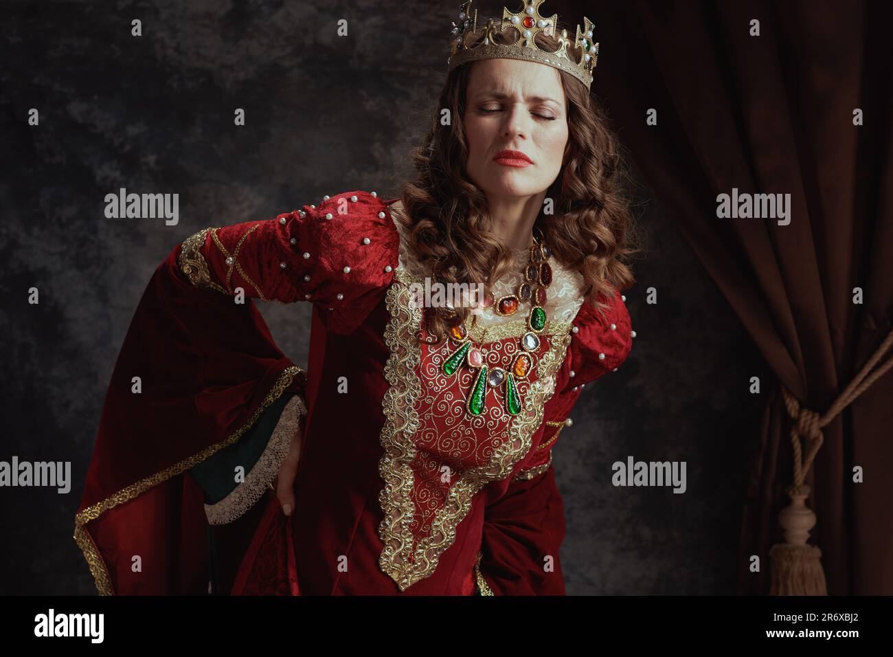 tired medieval queen in red dress with crown having backache on dark ...
