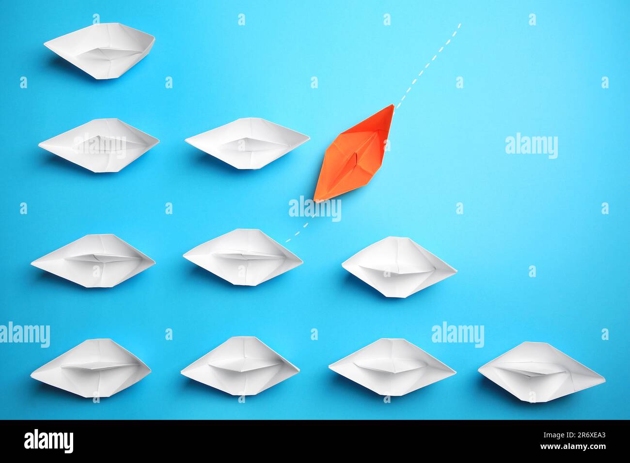 Orange paper boat floating away from others on light blue background ...