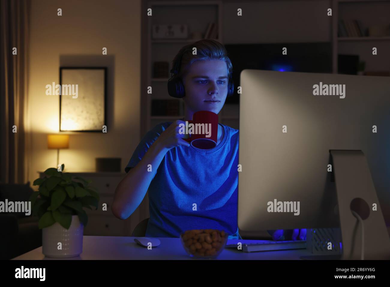 Teenage boy with cup of drink using computer in room at night. Internet ...