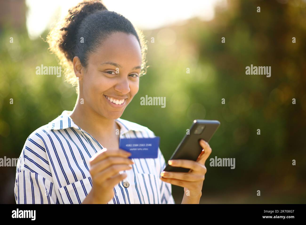 Black woman looking at you holding credit card and phone in a park Stock Photo - Alamy
