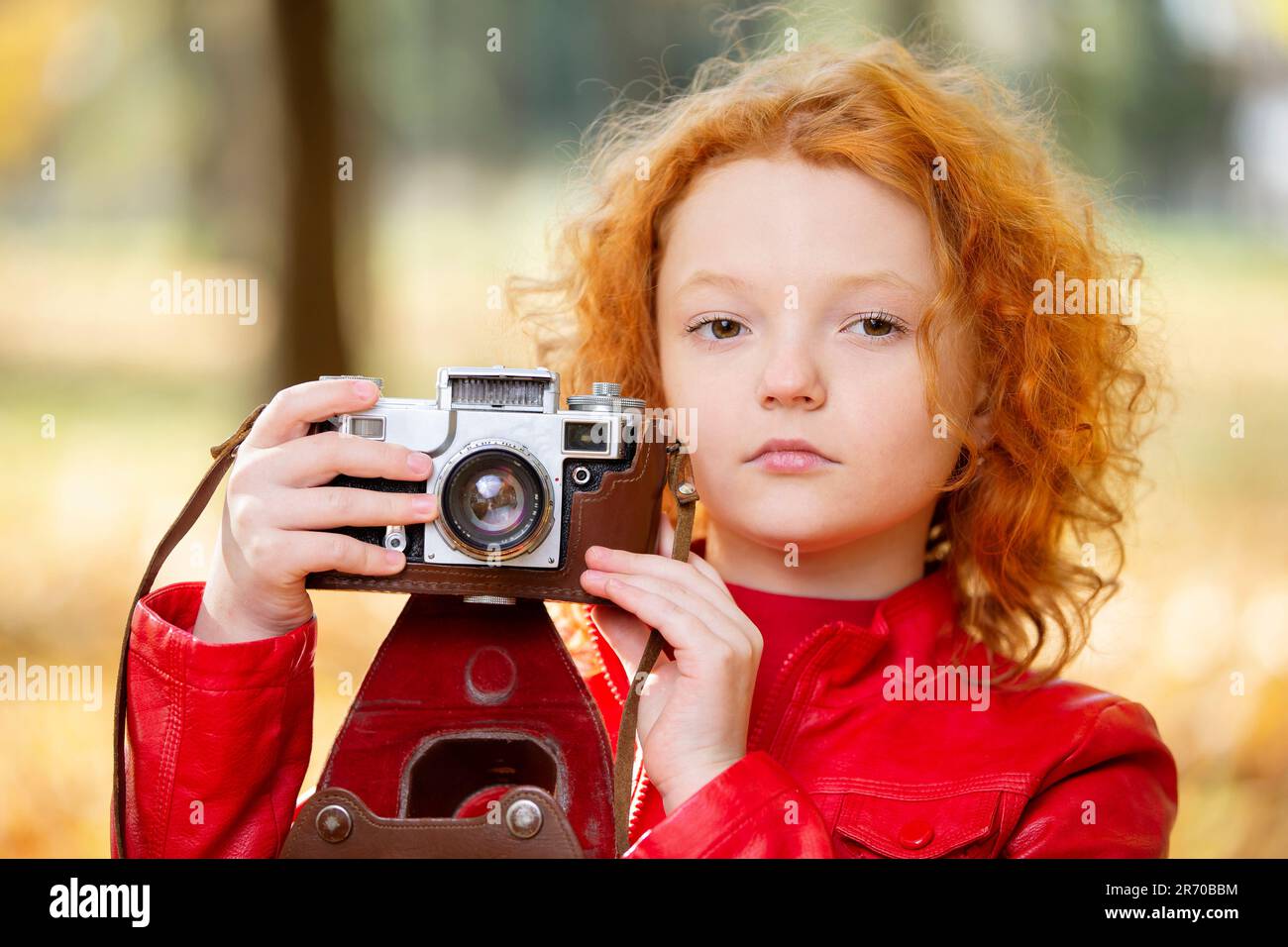 Little red-haired girl with a retro camera on an autumn background.Little red-haired girl with a ...