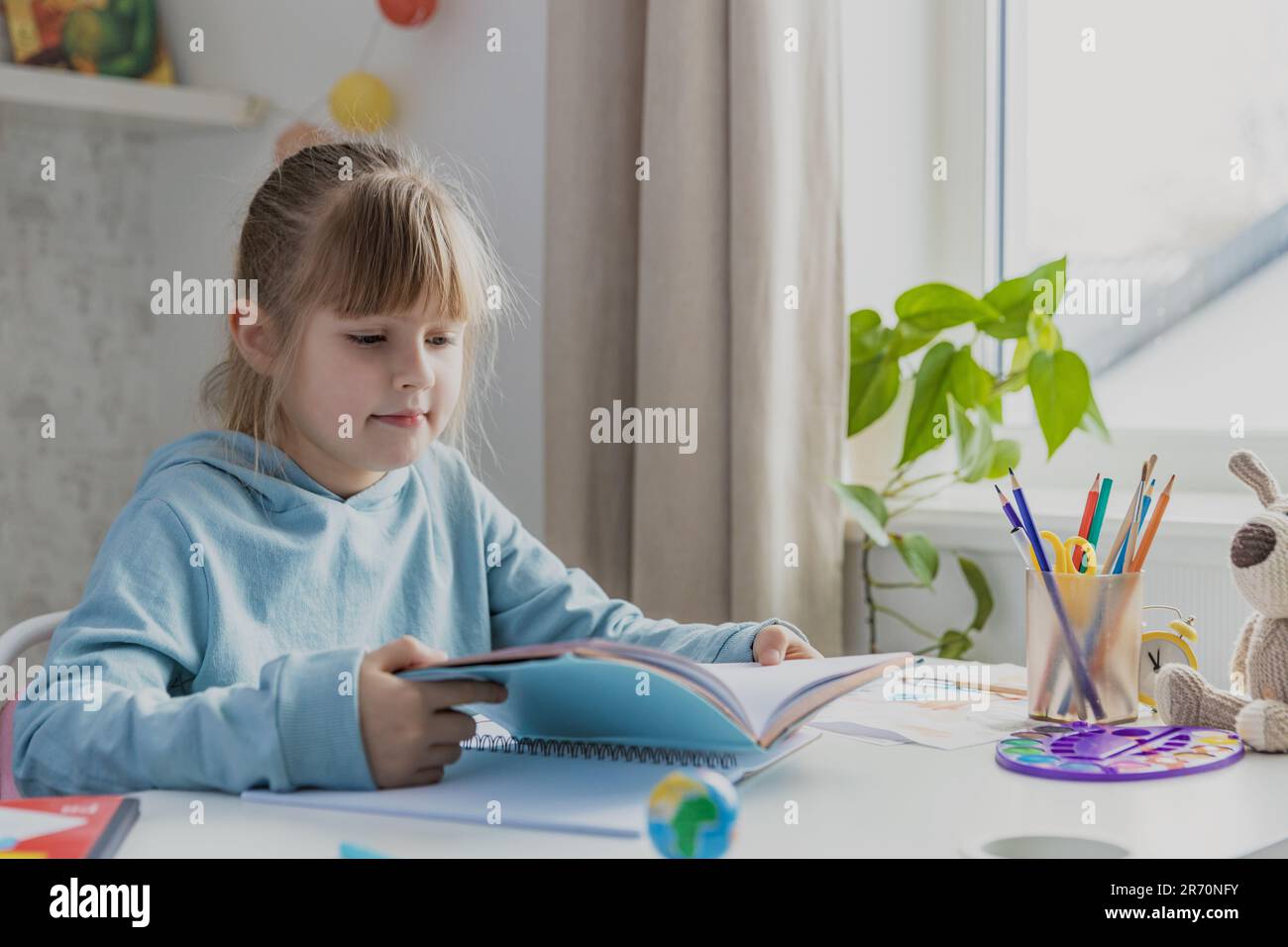 Schooler cute little girl sitting at desk in bedroom, holding a book and reading literature ...