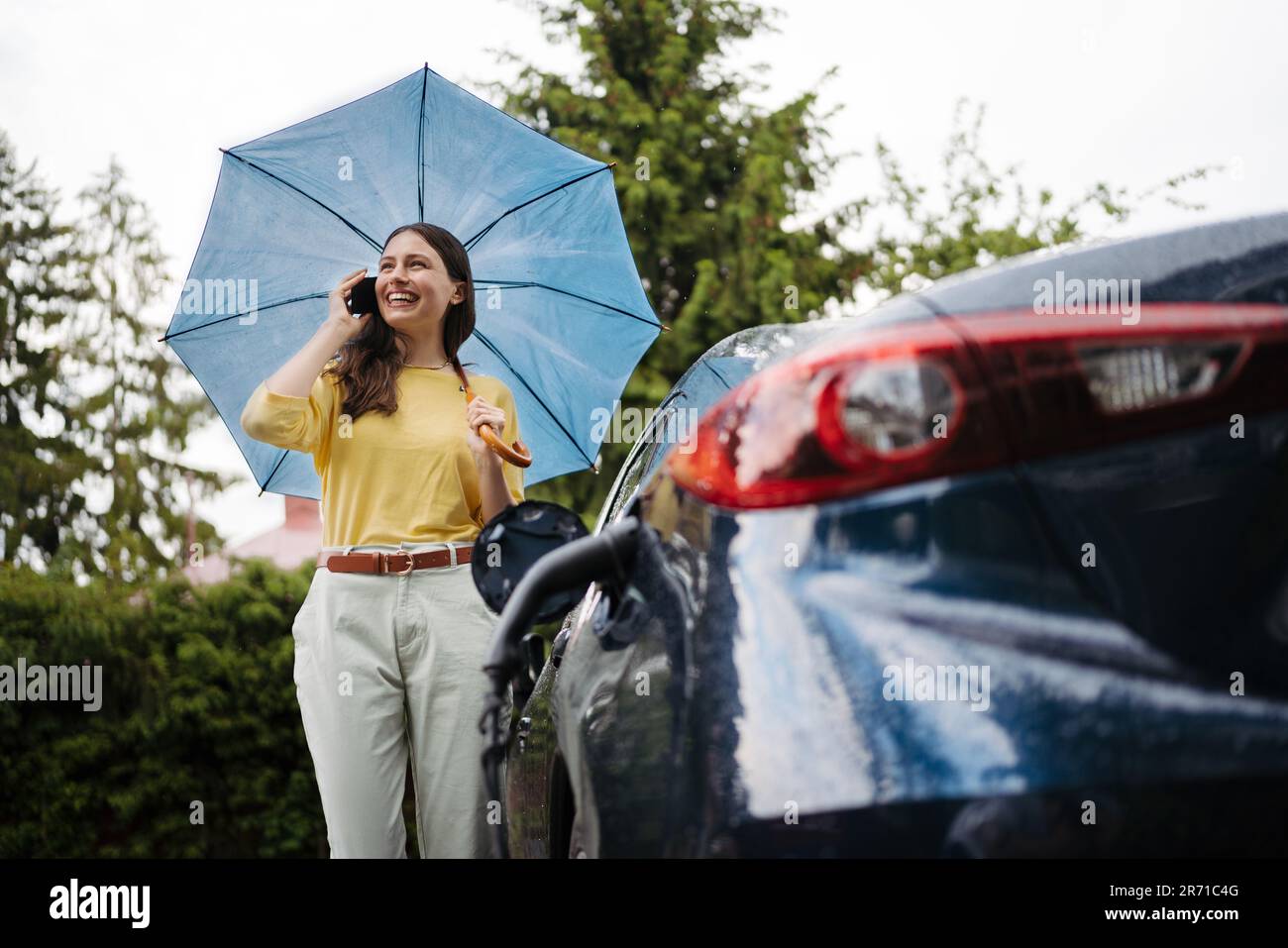 Woman calling, standing under umbrella while charging her electric car ...