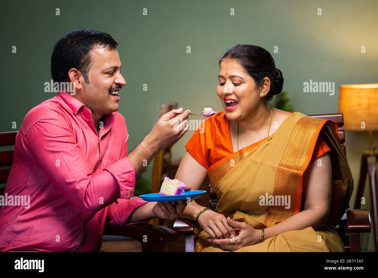 Happy husband feeding cake to wife while sitting on sofa at home ...