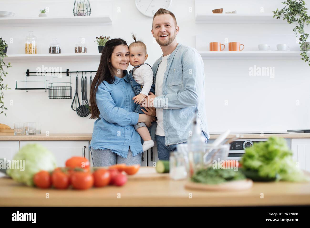 Portrait of joyful young parents in denim clothes cuddling little ...