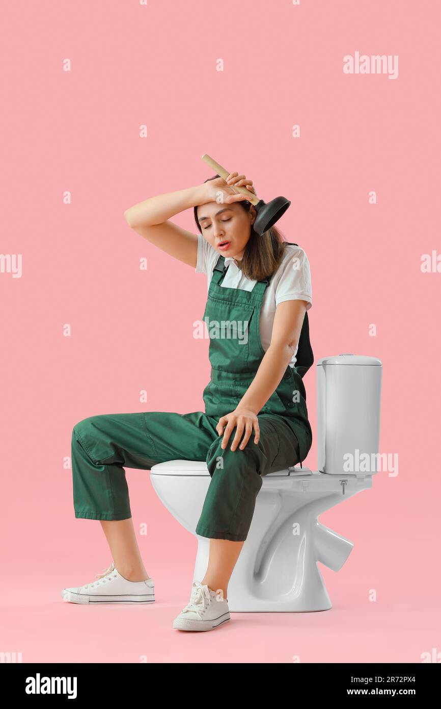 Tired female plumber with plunger sitting on toilet bowl against pink ...