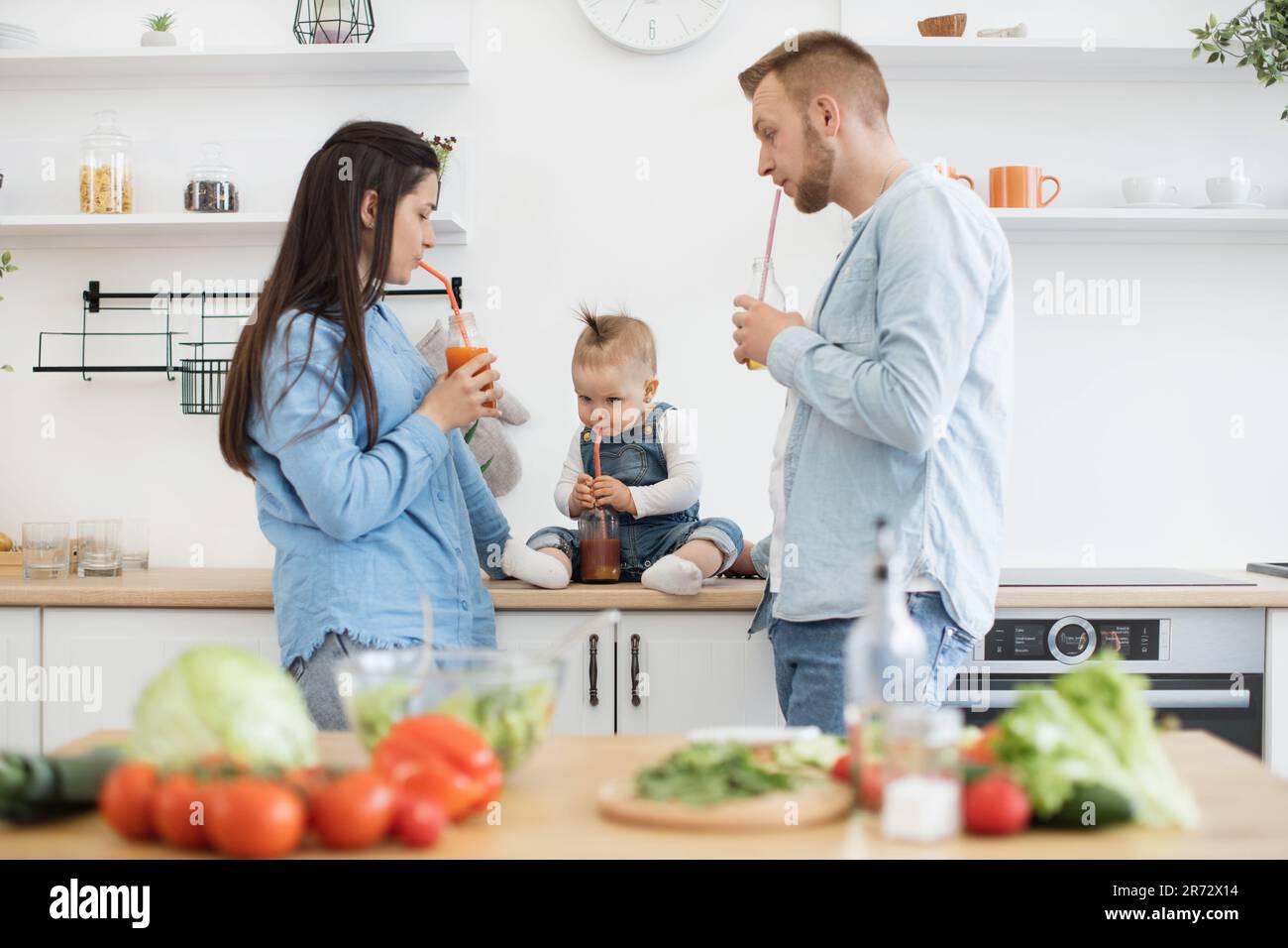 Delightful parents and kid enjoying refreshing glass of fresh juice while creating meal in ...