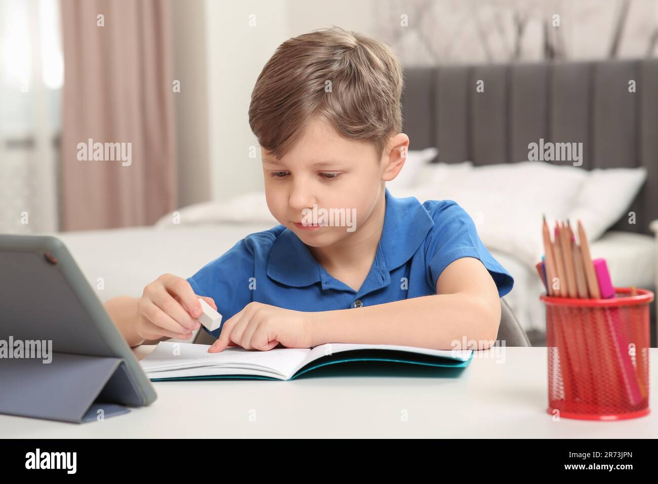 Little boy erasing mistake in his notebook near tablet at white desk ...