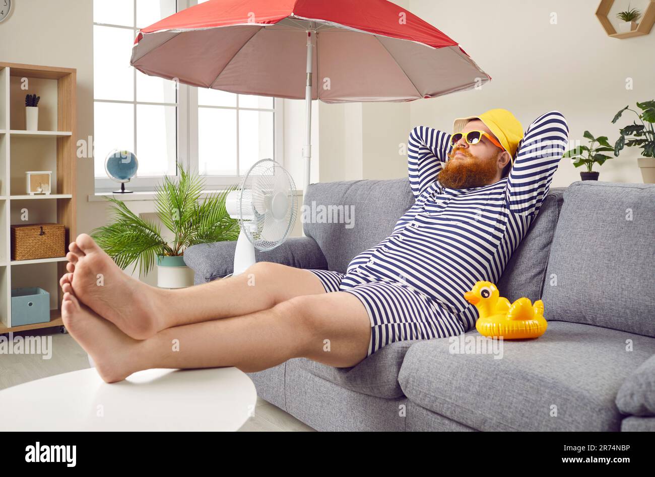 Funny fat man spends summer vacation at home sitting on sofa under fan ...