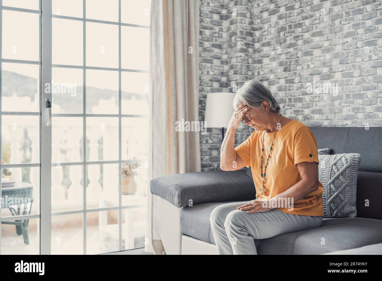 Sad depressed middle aged mature woman sitting alone at home touching head feeling headache ...