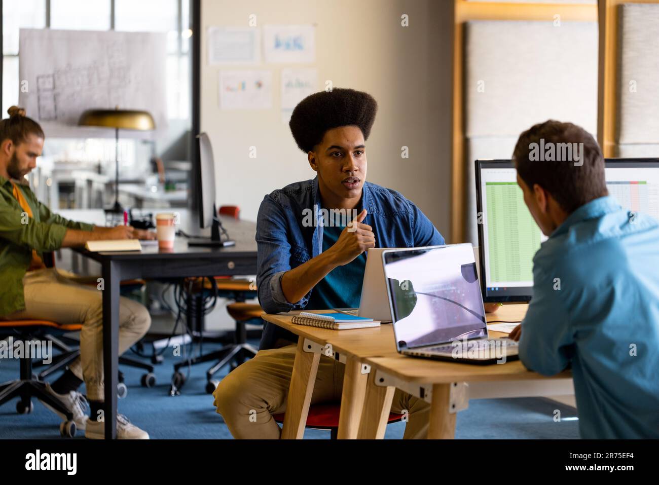 Diverse male colleagues in discussion at desks using laptops in office ...
