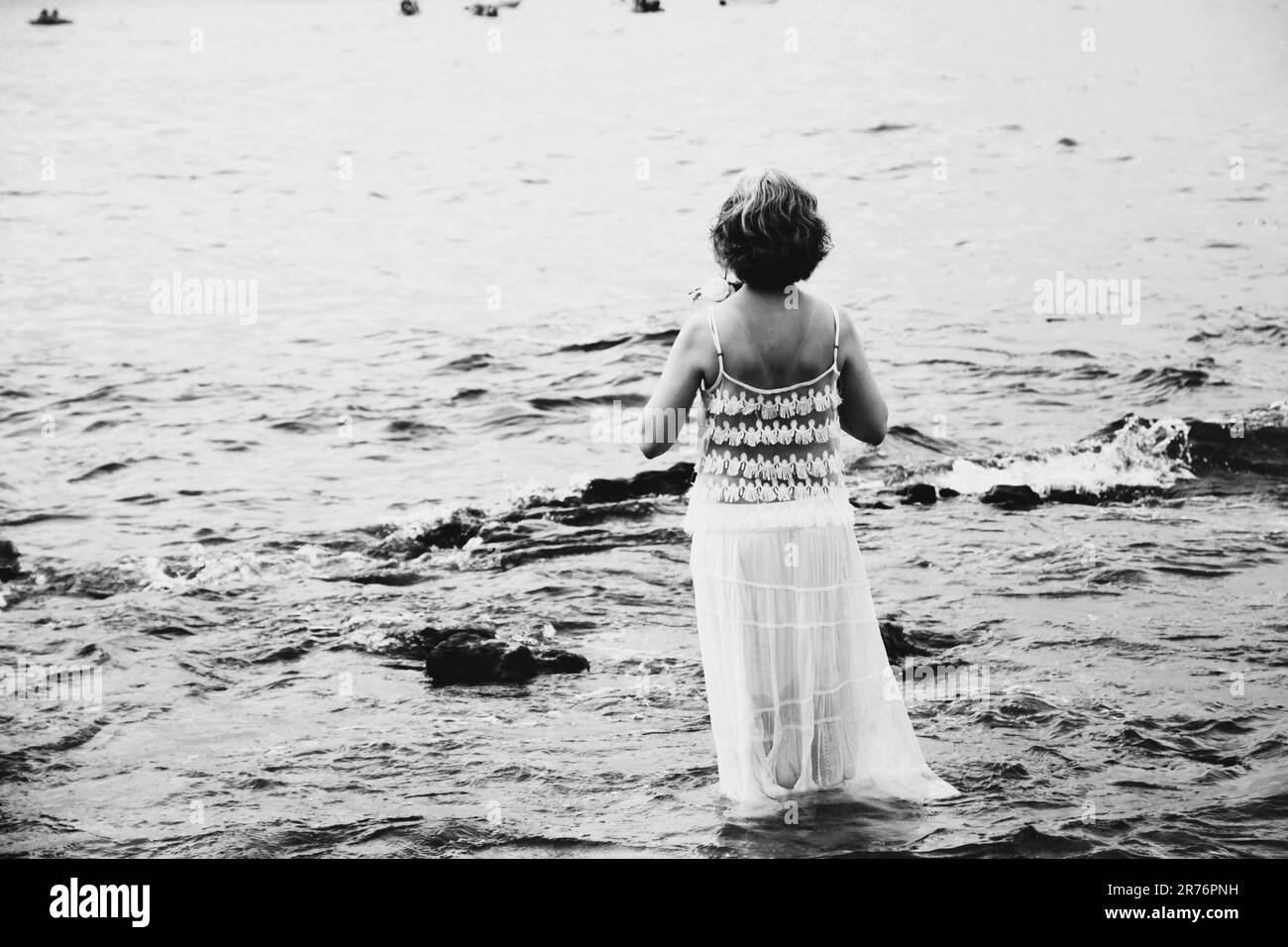 A young female child wearing a white dress is standing in shallow ocean ...
