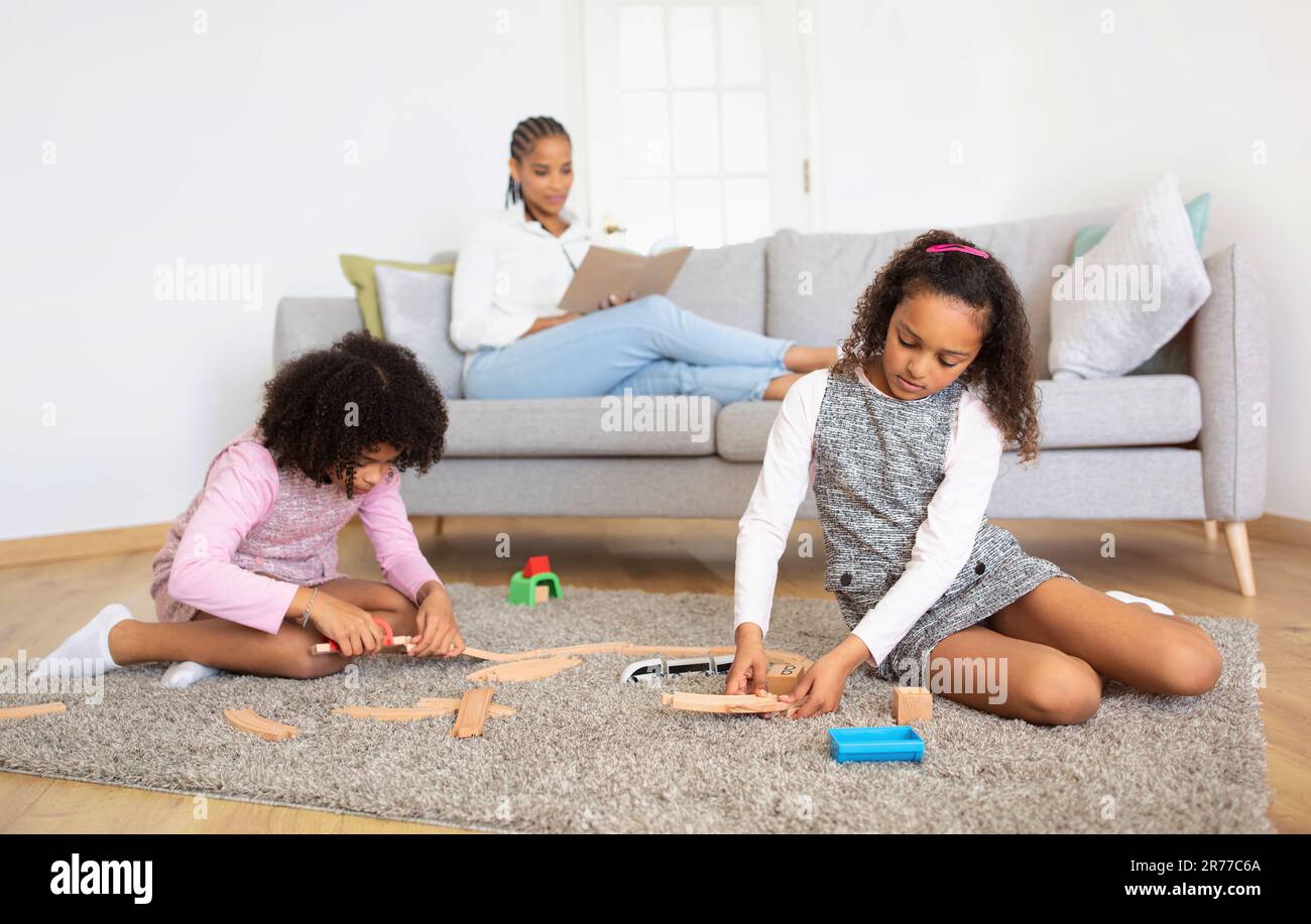 Black Daughters Playing With Toy Railway While Mother Reading Indoor Stock Photo - Alamy