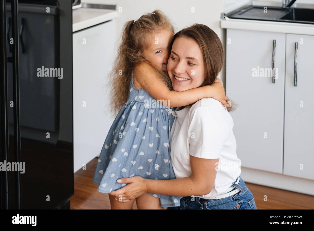 Child whispering something on Mother's ear at home on the kitchen. Kid ...